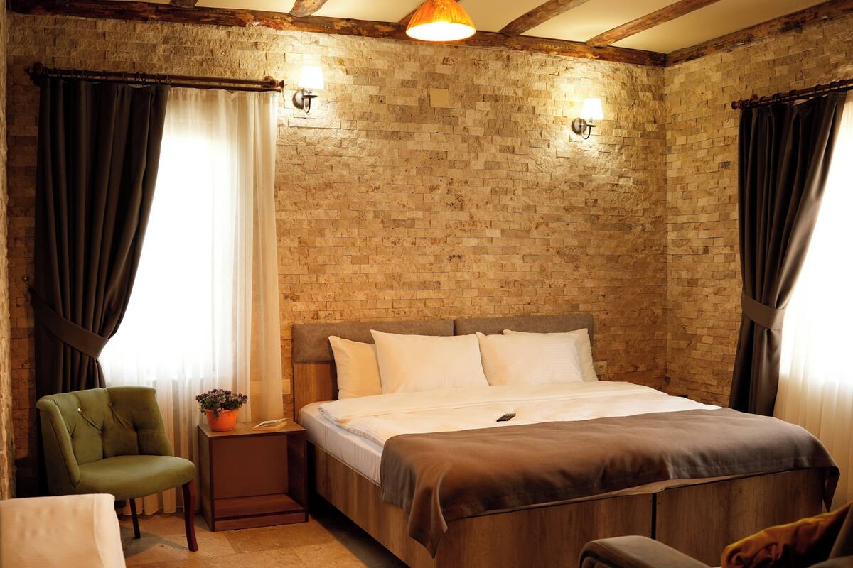 Quarto com parede de tijolos, cama com roupa de cama branca, poltrona verde e mesa de cabeceira com abajur.