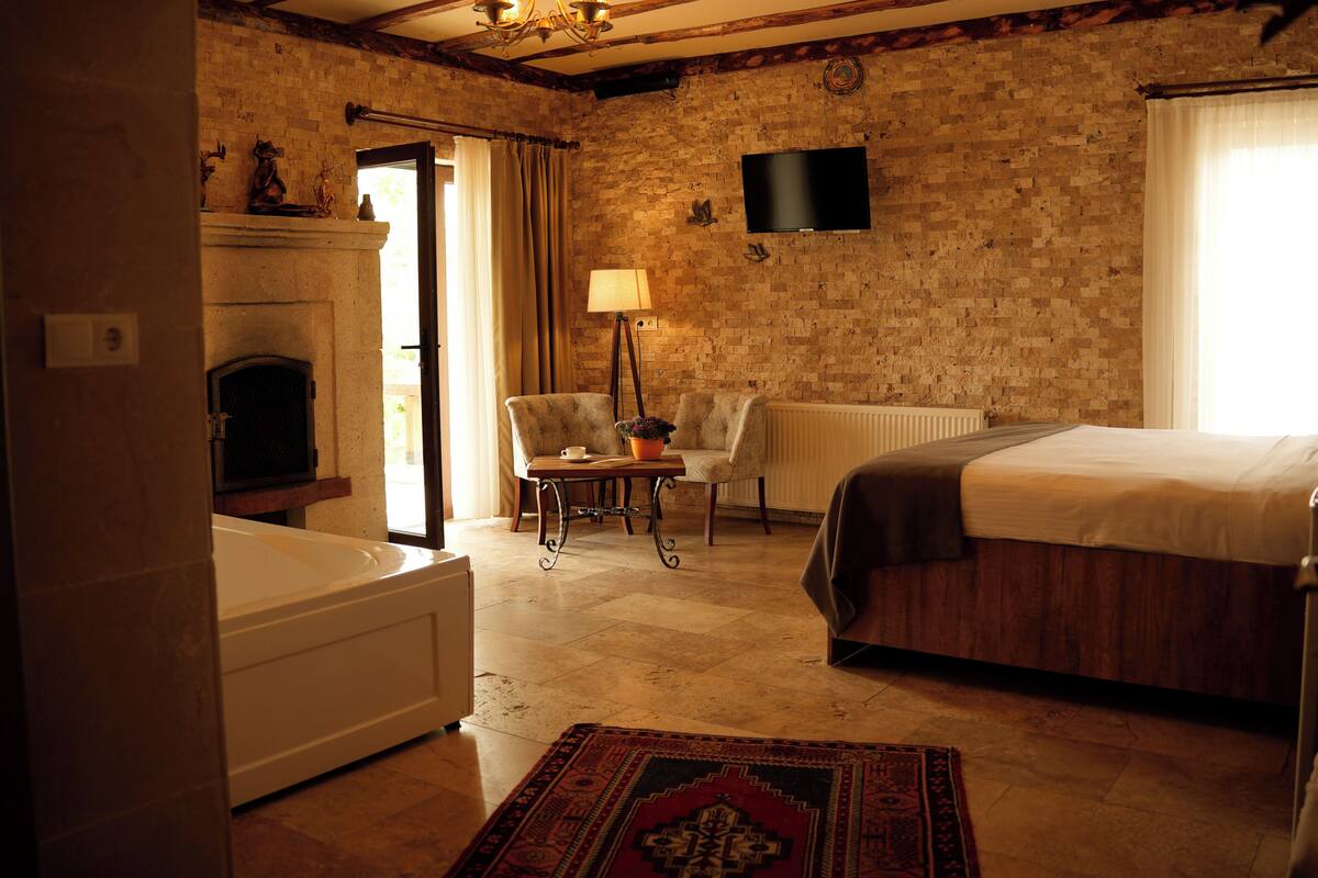 Quarto de hotel com parede de tijolos, lareira, cama, poltrona, mesa com abajur e TV fixada na parede.