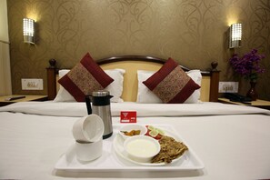 Cama bem arrumada com bandeja de comida, incluindo pão achatado, salada e uma xícara de café, em quarto de hotel com papel de parede estampado e arandelas.