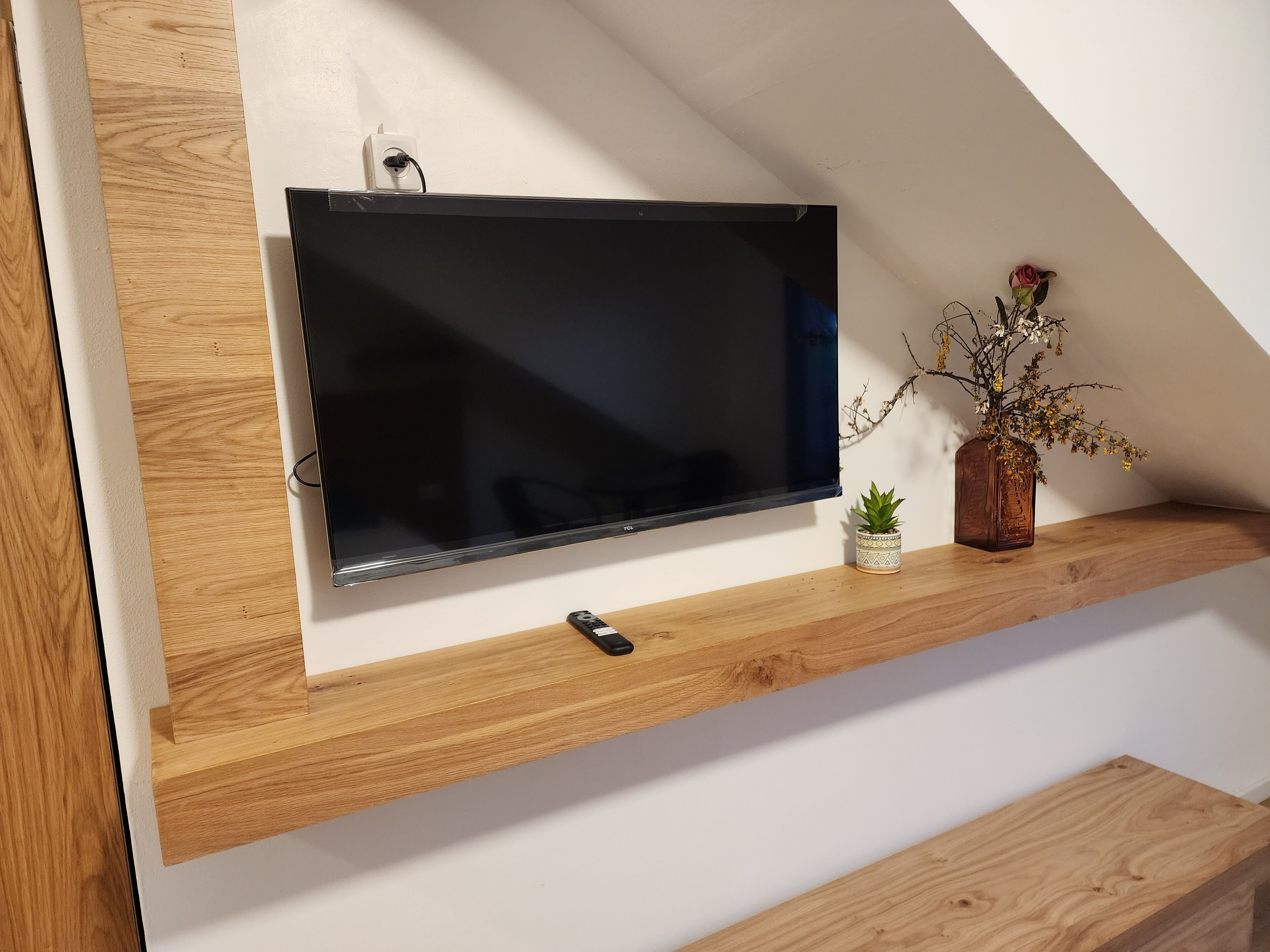 Uma TV de tela plana presa em uma prateleira de madeira, com uma tomada, um controle remoto e um pequeno vaso de planta.