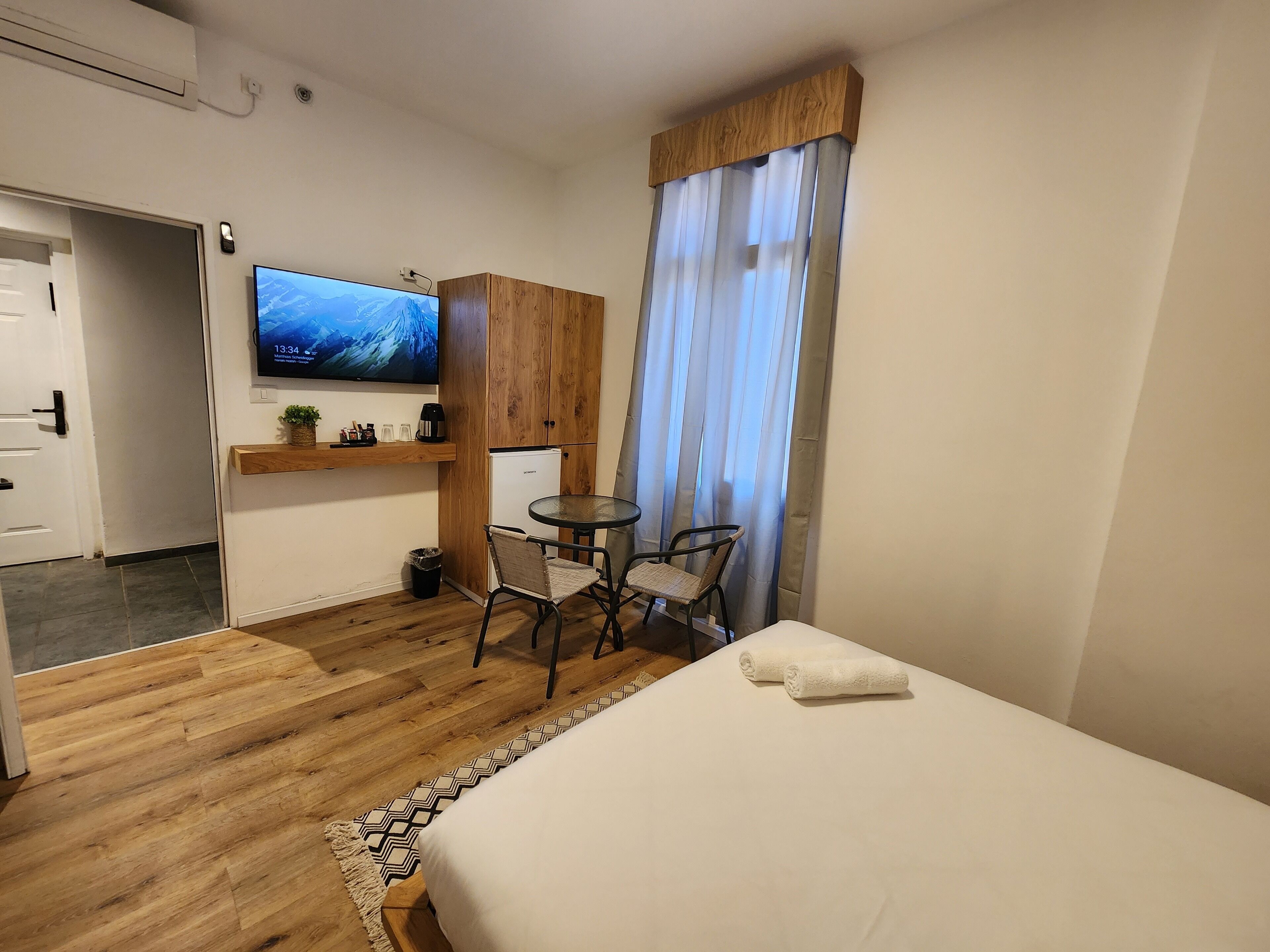 Quarto de hotel com cama, uma pequena mesa com cadeiras, televisão, armário de madeira e janela com cortinas.
