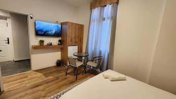 Quarto de hotel com cama, uma pequena mesa com cadeiras, televisão, armário de madeira e janela com cortinas.