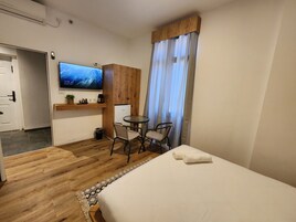 Quarto de hotel com cama, uma pequena mesa com cadeiras, televisão, armário de madeira e janela com cortinas.