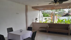 Uma área de refeições com mesa, cadeiras, ventilador de teto e vista para o verde lá fora.