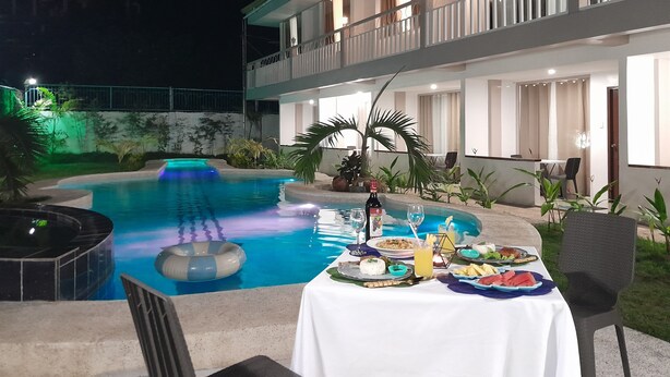 Uma área de refeições à beira da piscina, com uma mesa posta para refeição, cercada por uma piscina e um edifício com varanda.