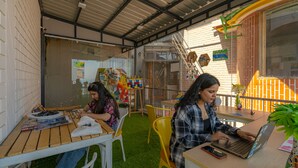 Um café ao ar livre com mesas e cadeiras de madeira, piso de grama sintética verde e uma parede de tijolos com uma janela.