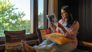 Uma pessoa sentada num banco lendo um livro, com uma janela ao fundo.