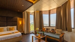 Quarto de hotel moderno com uma cama grande, um sofá, uma mesa de centro de madeira e vista para as montanhas.