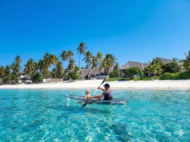 Um casal em um barco pequeno em águas turquesa cristalinas, com uma praia e palmeiras ao fundo.