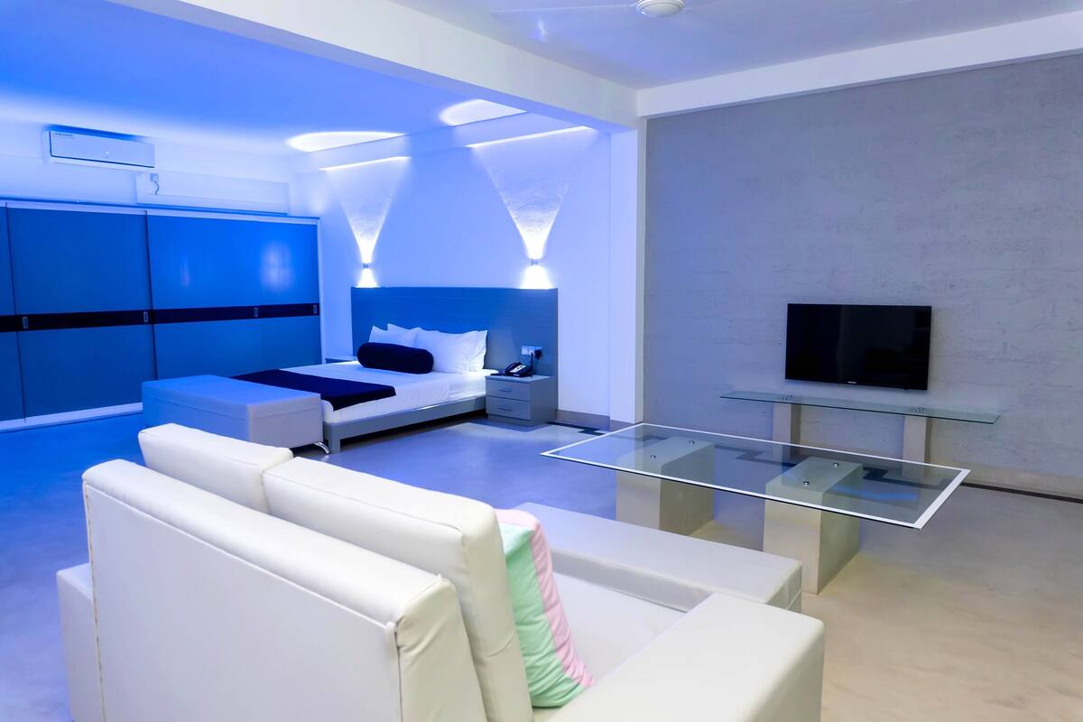 Quarto de hotel moderno com uma cama, sofás brancos, uma mesa de centro de vidro e uma televisão de tela plana fixada na parede.