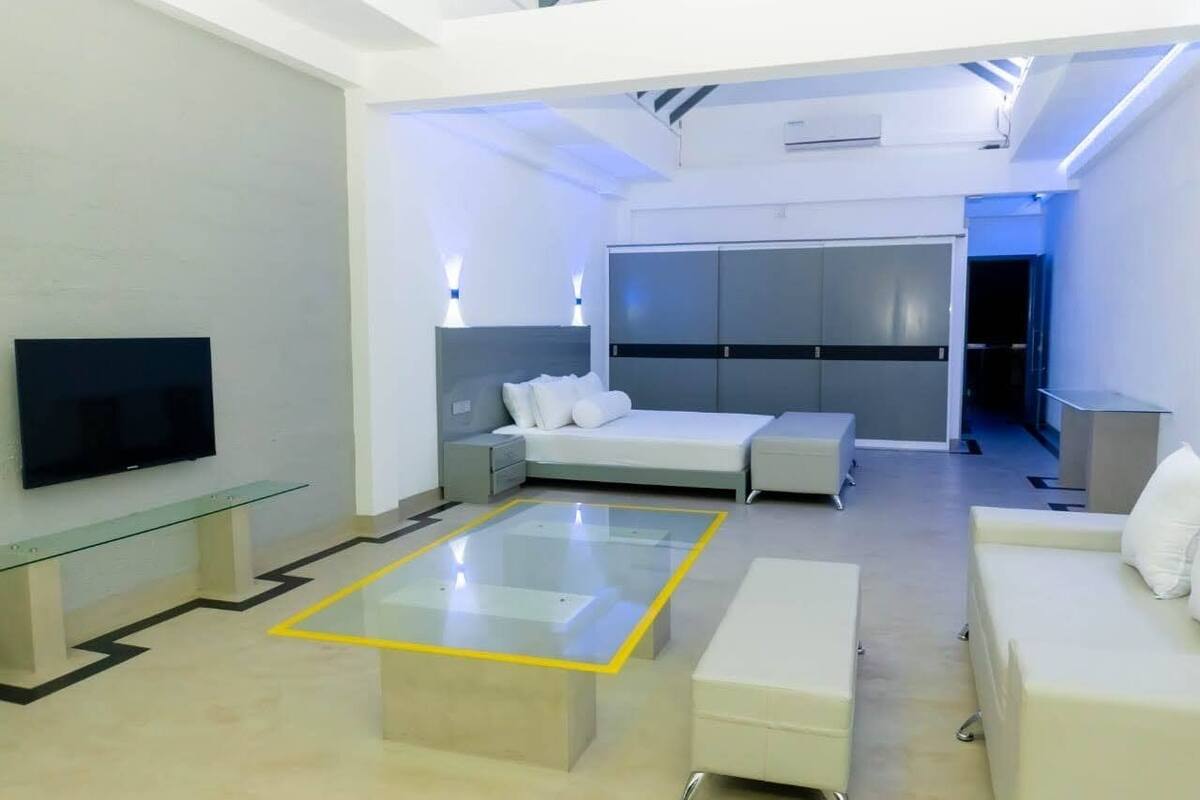 Quarto de hotel moderno com uma cama, uma televisão, uma mesa de centro de vidro e um sofá.