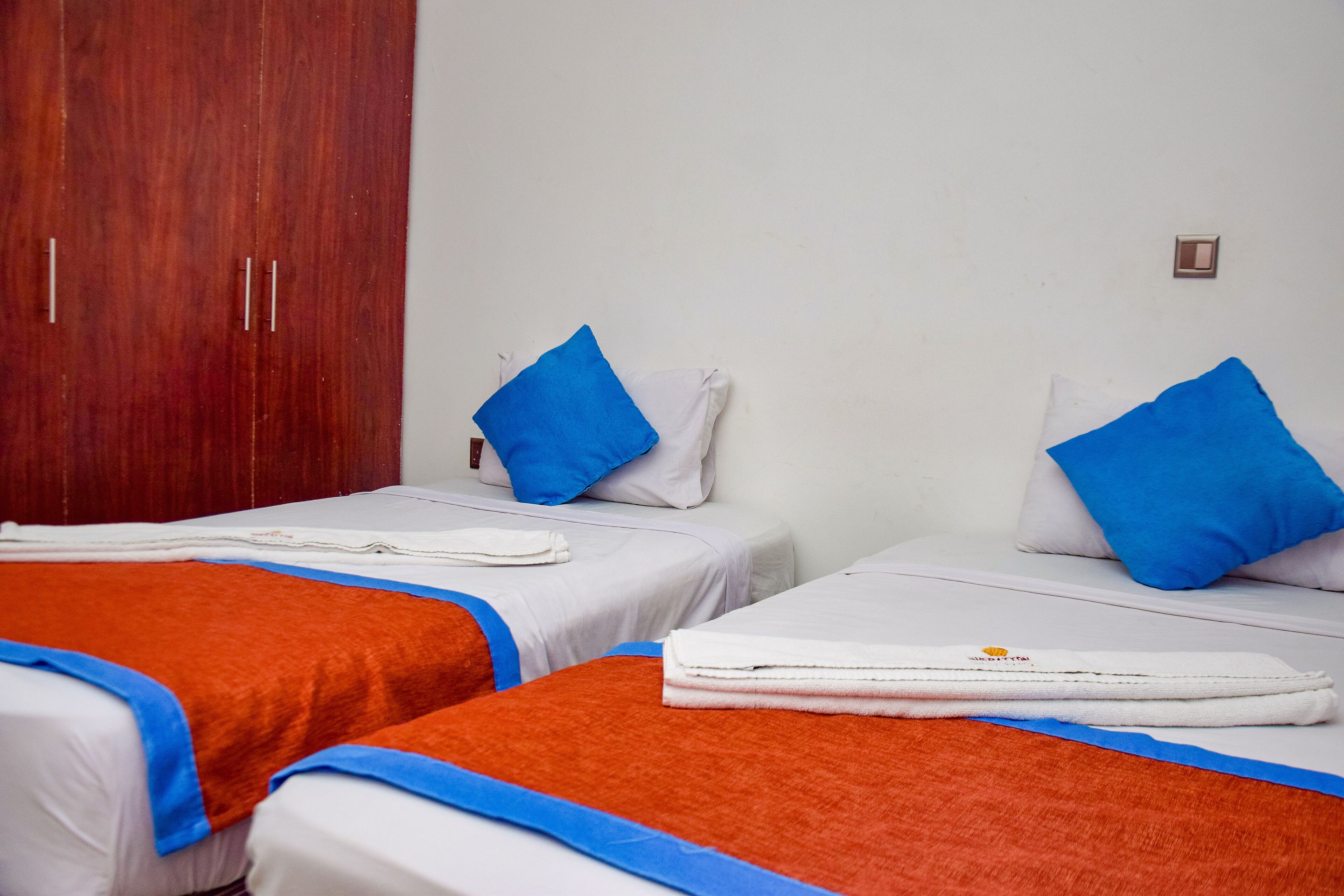 Quarto de hotel com duas camas, cada uma com colcha laranja e azul, roupa de cama branca e almofadas azuis.