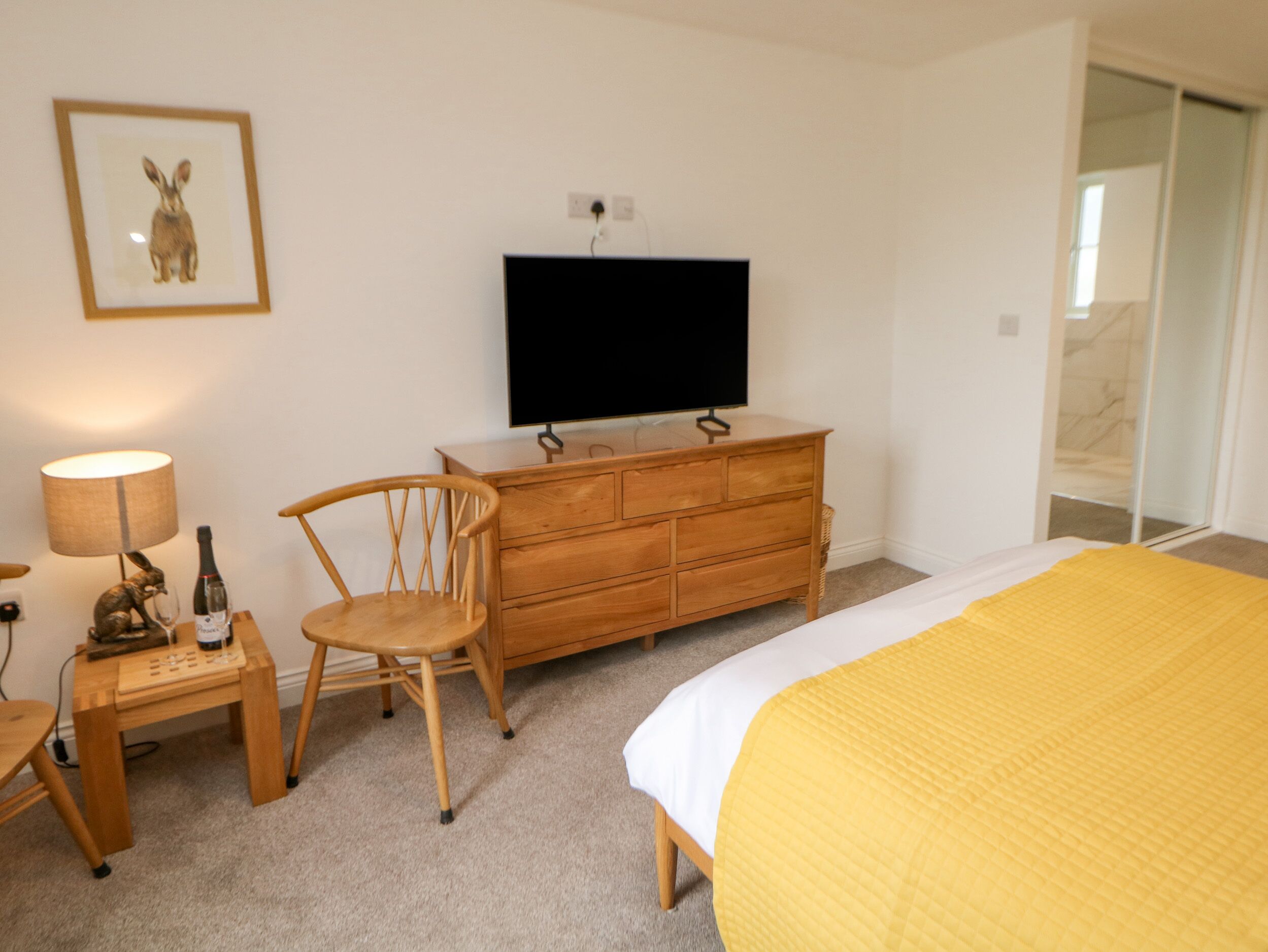 Quarto com cama, cómoda de madeira, televisão, cadeira e pequena mesa com abajur e garrafa.