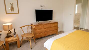 Quarto com cama, cómoda de madeira, televisão, cadeira e pequena mesa com abajur e garrafa.
