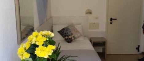 Quarto com cama, mesa de cabeceira e espelho. Há uma porta e uma pequena planta na mesa de cabeceira.