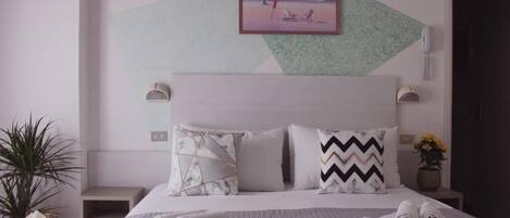 Uma cama bem arrumada com almofadas, uma mesinha de cabeceira, um relógio de parede e um quadro emoldurado.
