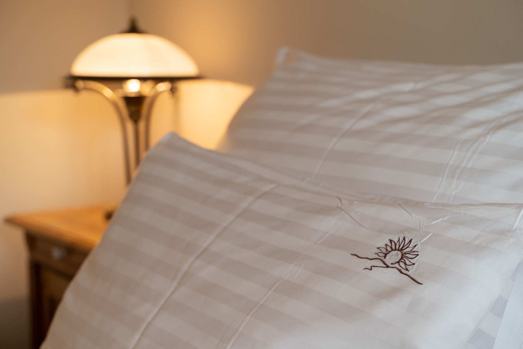 Cama com almofadas brancas e um pequeno desenho bordado de sol, luminária de cabeceira e mesa de cabeceira de madeira.