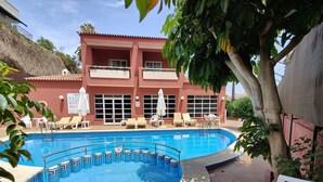 Uma piscina com design curvo, rodeada por espreguiçadeiras e guarda-sóis, com um edifício com varandas e janelas ao fundo.