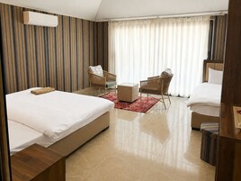 Quarto de hotel com duas camas, um espaço de estar com uma poltrona e apoio para os pés, uma janela grande com cortinas e um ar condicionado fixado na parede.