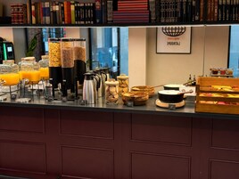 Um buffet de café da manhã com diversos alimentos e bebidas, uma máquina de café e uma estante com livros.