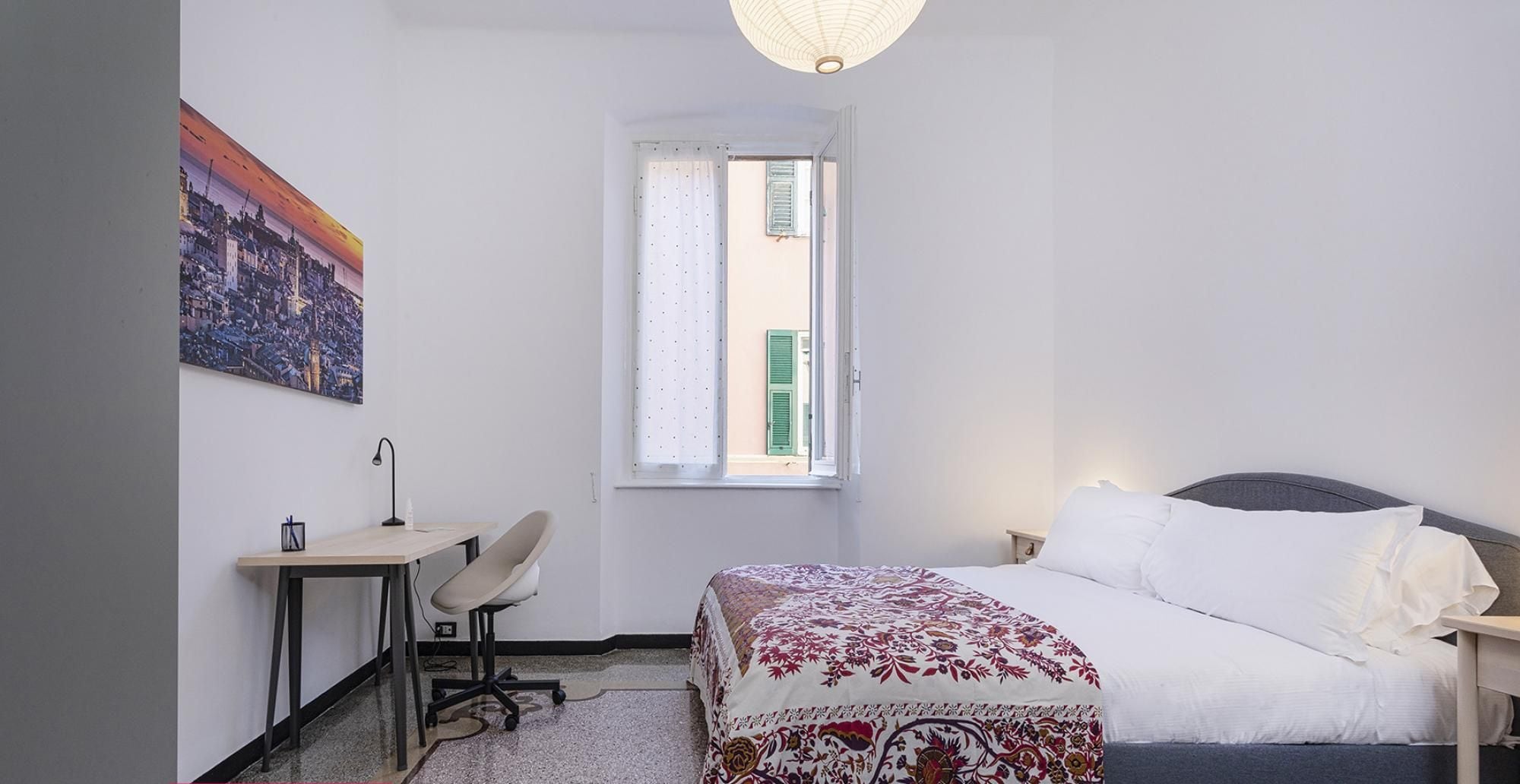 Um quarto com cama, escrivaninha, cadeira e janela.