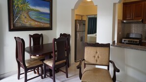 Uma área de jantar com uma mesa e cadeiras de madeira escura, um quadro representando uma cena de praia e uma cozinha com armários de madeira e um fogão.