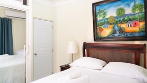 Quarto de hotel com uma cama, mesa de cabeceira com abajur, quadro na parede e porta que dá para outro cômodo.