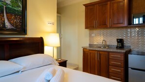 Quarto de hotel com cama, mesa de cabeceira, luminária, pintura, armários de madeira e uma cozinha equipada com pia, cafeteira e geladeira.
