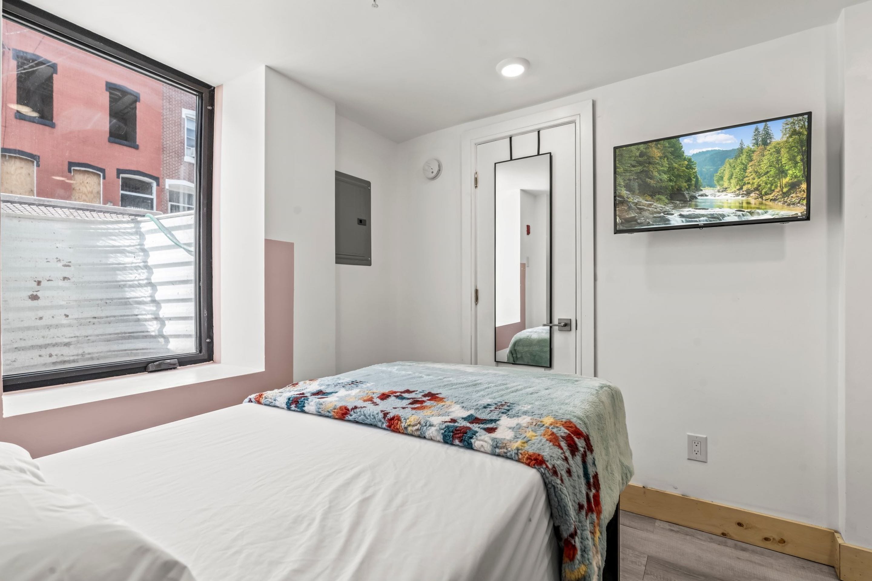 Quarto com cama, janela com vista para edifícios, TV na parede e porta com espelho.
