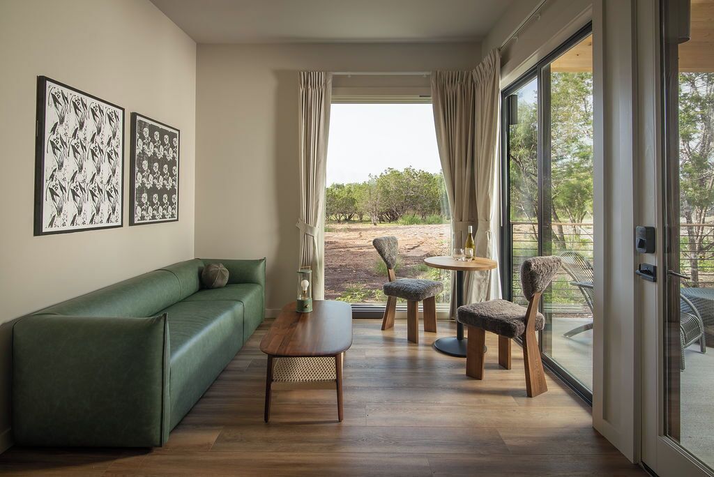 Uma sala de estar moderna com um sofá verde, uma mesa de centro de madeira e duas poltronas. Há duas obras de arte emolduradas na parede e uma janela grande com cortinas.