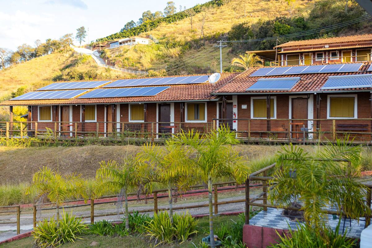 Um edifício com painéis solares no telhado, uma escadaria que sobe uma colina e uma área cercada com plantas.