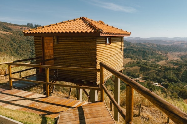 Uma cabana de madeira com telhado de cerâmica, localizada sobre uma plataforma de madeira com guarda-corpo, com vista para uma paisagem ondulada.