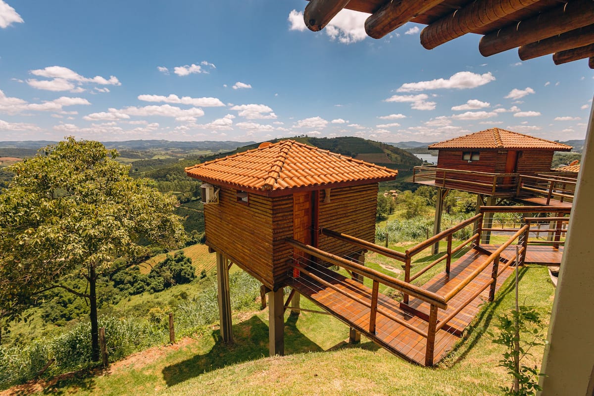 Duas cabanas de madeira com telhados de terracota, erguidas em estacas, ligadas por passarelas de madeira, com vista para colinas verdejantes e um céu azul claro.