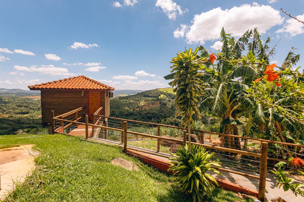 Uma cabana de madeira com telhado de telhas vermelhas, rodeada por vegetação exuberante e uma vista panorâmica de colinas ondulantes.