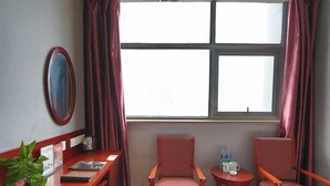 Quarto de hotel com uma escrivaninha de madeira, duas poltronas, uma mesinha com uma garrafa de água e uma janela grande com cortinas.