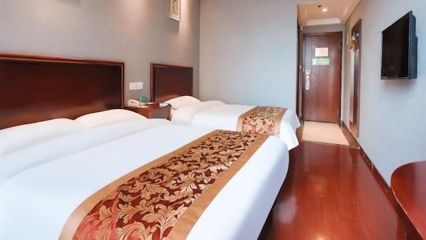 Quarto de hotel com duas camas, cabeceira de madeira, mesa de cabeceira com abajur e televisão fixada na parede.