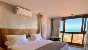 Quarto de hotel com uma cama grande, cabeceira de madeira, vista para o mar e um aparelho de ar condicionado na parede.