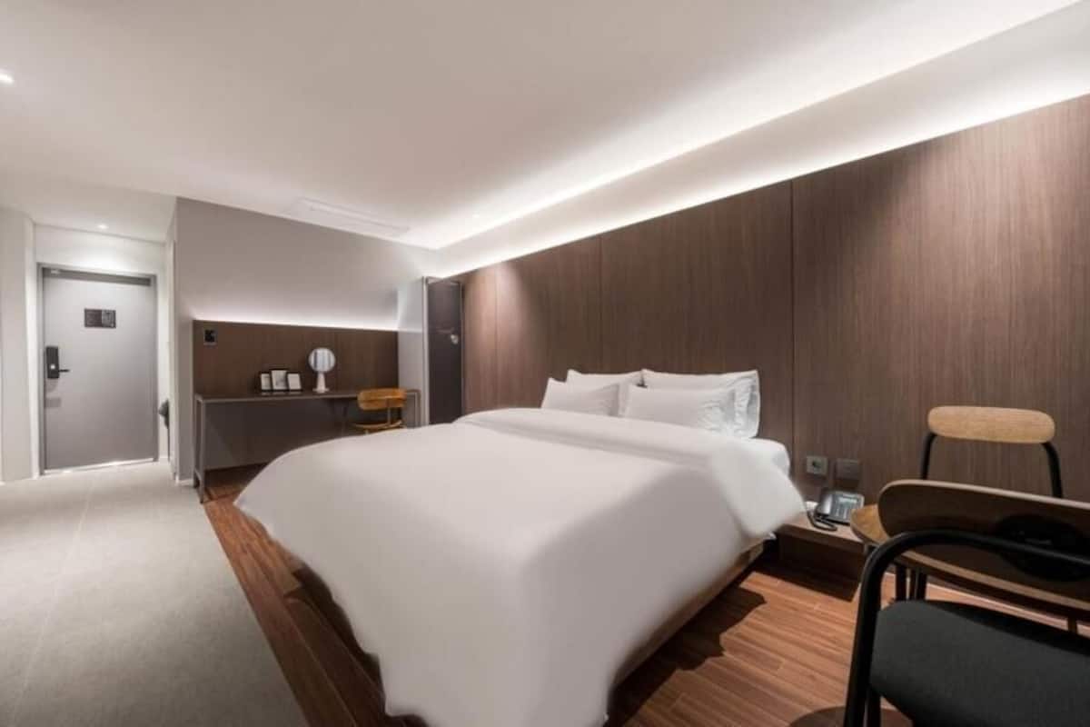 Quarto de hotel moderno com uma cama grande, uma mesa, uma cadeira e uma pequena mesa com abajur.