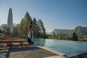 Uma pessoa caminhando sobre um deck de madeira, próximo a uma piscina, com vista para árvores e colinas.