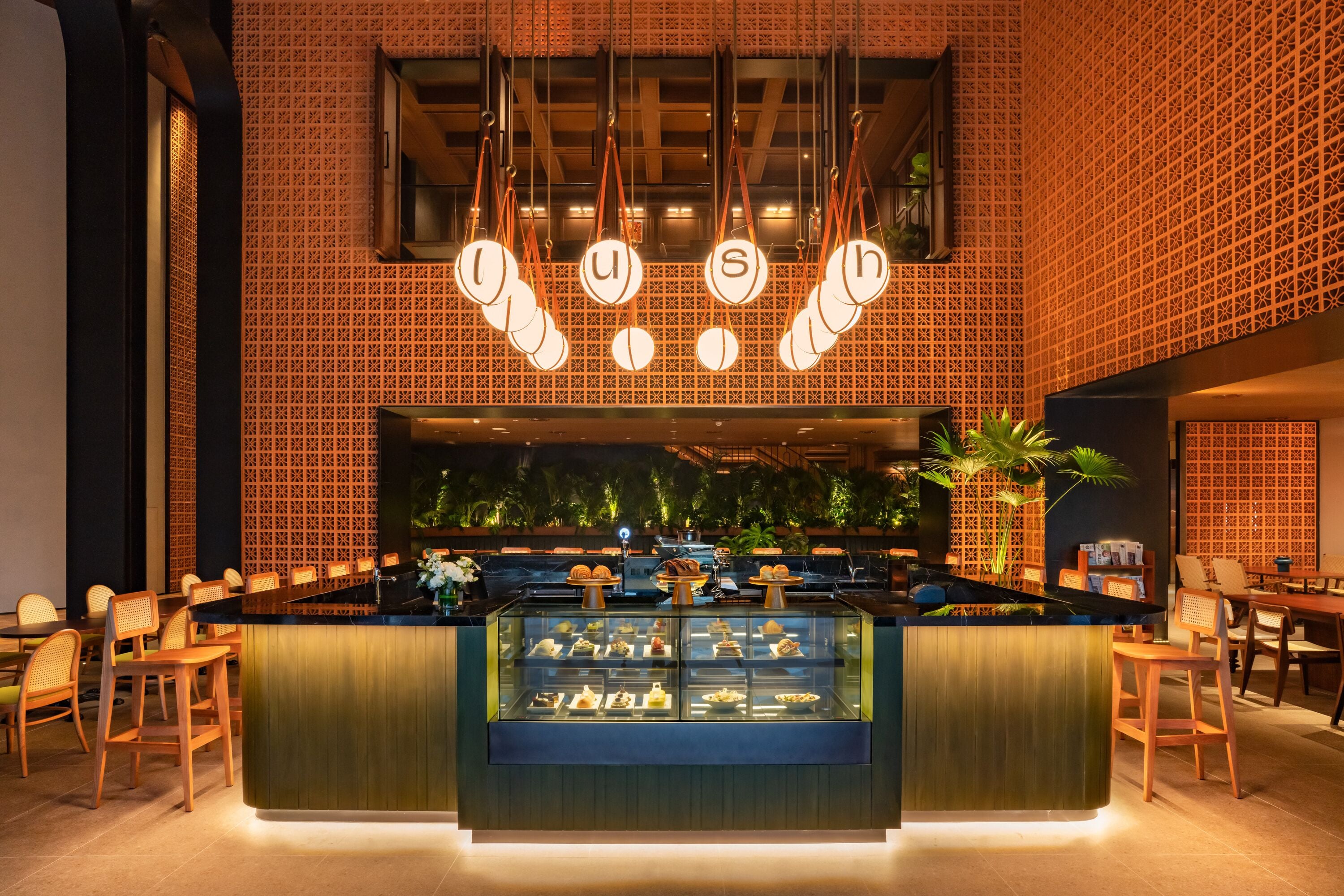 Uma área de bar moderna com um balcão central exibindo sobremesas, rodeada por cadeiras de madeira e uma luminária decorativa no teto.