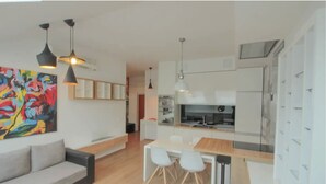 Uma sala de estar moderna com um sofá cinza, uma área de jantar com duas cadeiras brancas e uma cozinha com armários brancos e uma pia de aço inoxidável.
