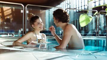 Um casal em uma piscina com uma mesa de jantar, cercado por grandes janelas e plantas dentro de casa.