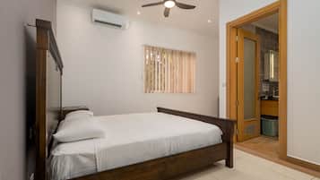 Quarto com cama de madeira, ventilador de teto, janela com persianas e porta que dá para um banheiro.