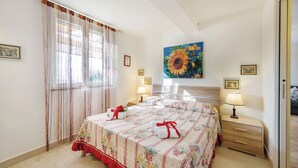 Quarto de hotel com cama, duas mesas de cabeceira, janela com cortinas e um quadro de girassóis na parede.