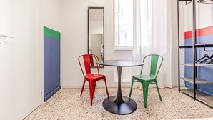 Quarto com uma mesa redonda, duas cadeiras, um espelho, uma janela com cortinas e um cabideiro.