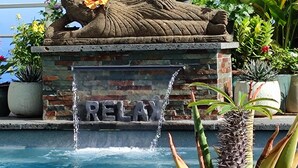 Uma área de piscina com uma estrutura de pedra exibindo a placa 'RELAX', cercada por plantas diversas e um céu azul límpido.