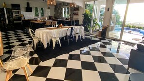 Uma sala de jantar com chão quadriculado, uma mesa posta para quatro pessoas e uma cozinha ao fundo.