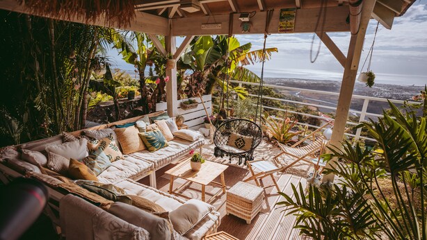Área de estar coberta ao ar livre com sofá, cadeiras e mesas, cercada por plantas tropicais e com vista panorâmica.