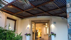 Entrada de um hotel com varanda coberta, plantas em vasos e balcão de recepção.