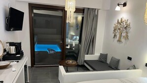 Quarto de hotel com uma cama, área de estar com almofadas, máquina de café, televisão e vista para uma piscina.